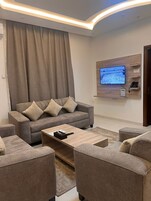 Uma sala de estar moderna com um sofá cinzento, poltronas a combinar e uma mesa de centro de madeira. Uma televisão de tela plana está fixada na parede, e um telefone está colocado numa prateleira.