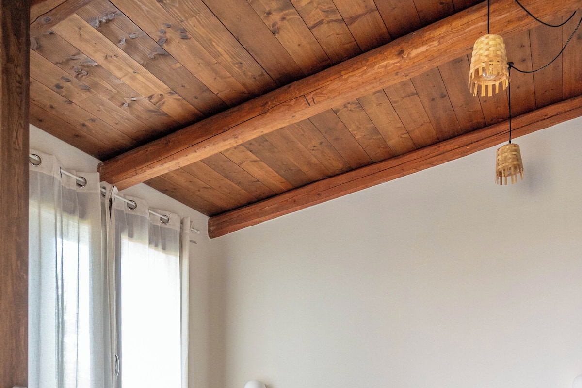 Quarto com teto de madeira, cama com cabeceira branca e luminária pendente.