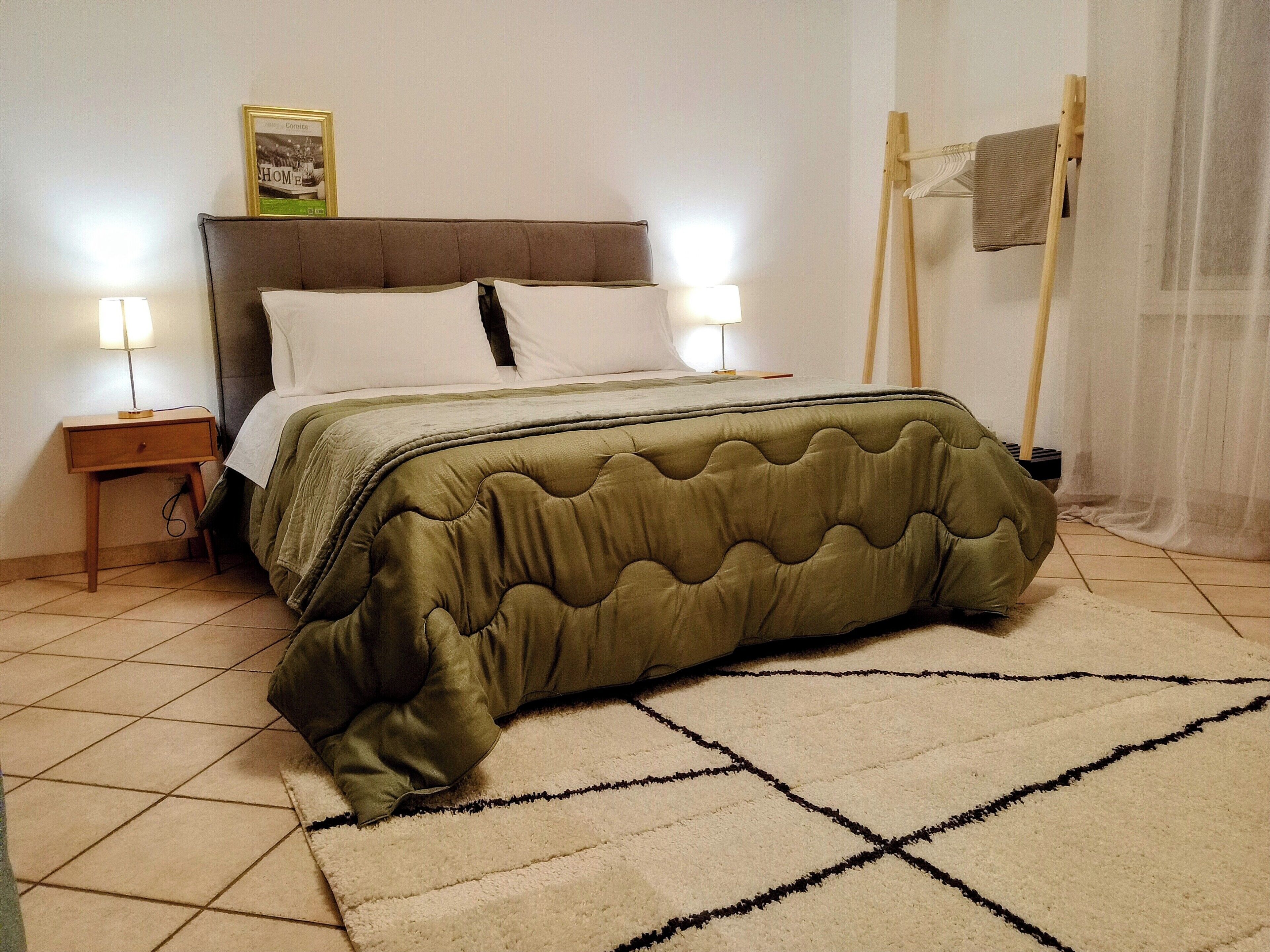 Cama bem arrumada com colcha verde e almofadas brancas, mesa de cabeceira com abajur e um quadro pendurado na parede.