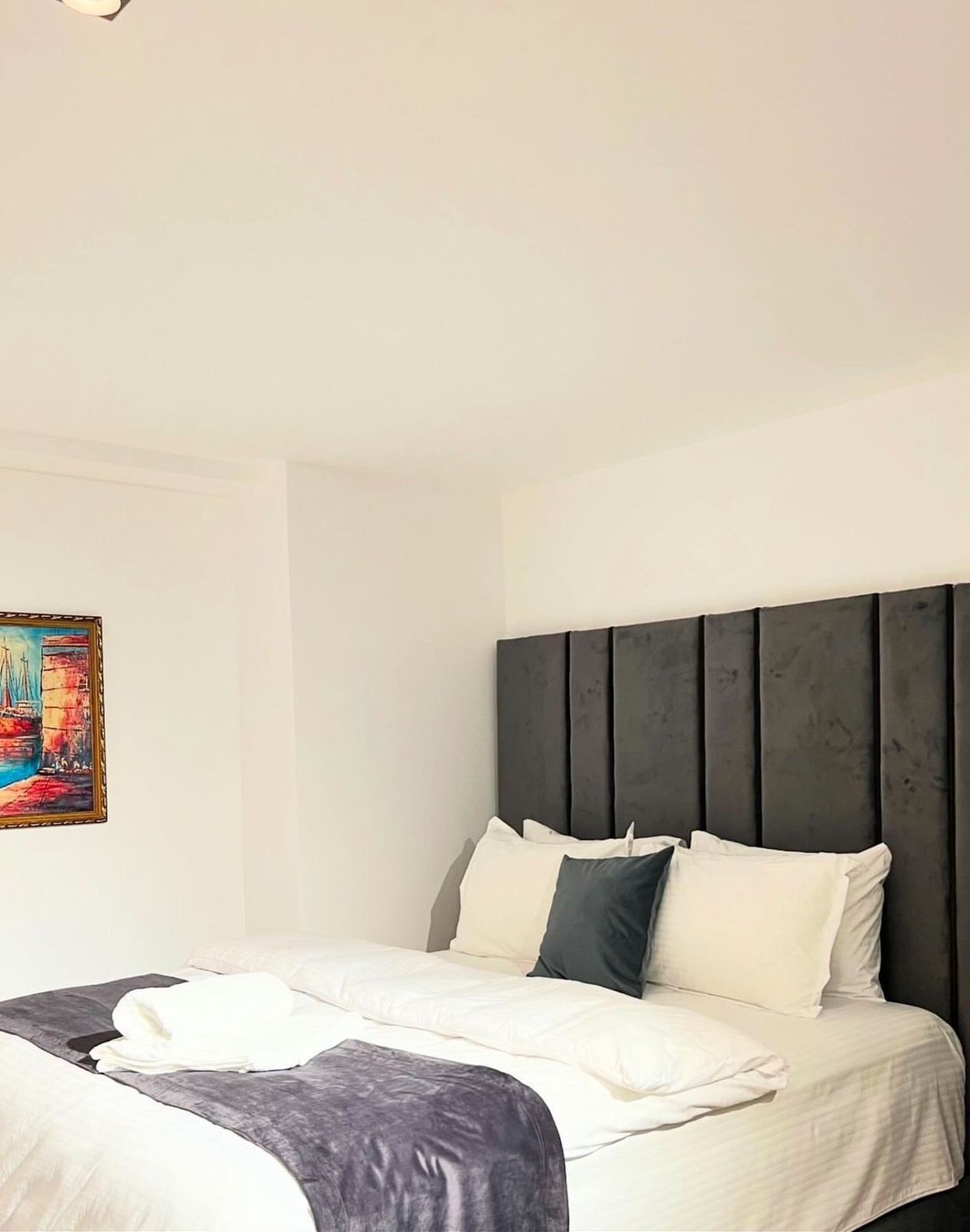 Cama bem arrumada com cabeceira cinza, roupa de cama branca e um quadro com vista urbana na parede.