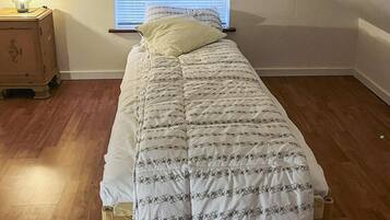 Cama de solteiro com colcha estampada em floral num quarto com piso de madeira e janela com persianas.