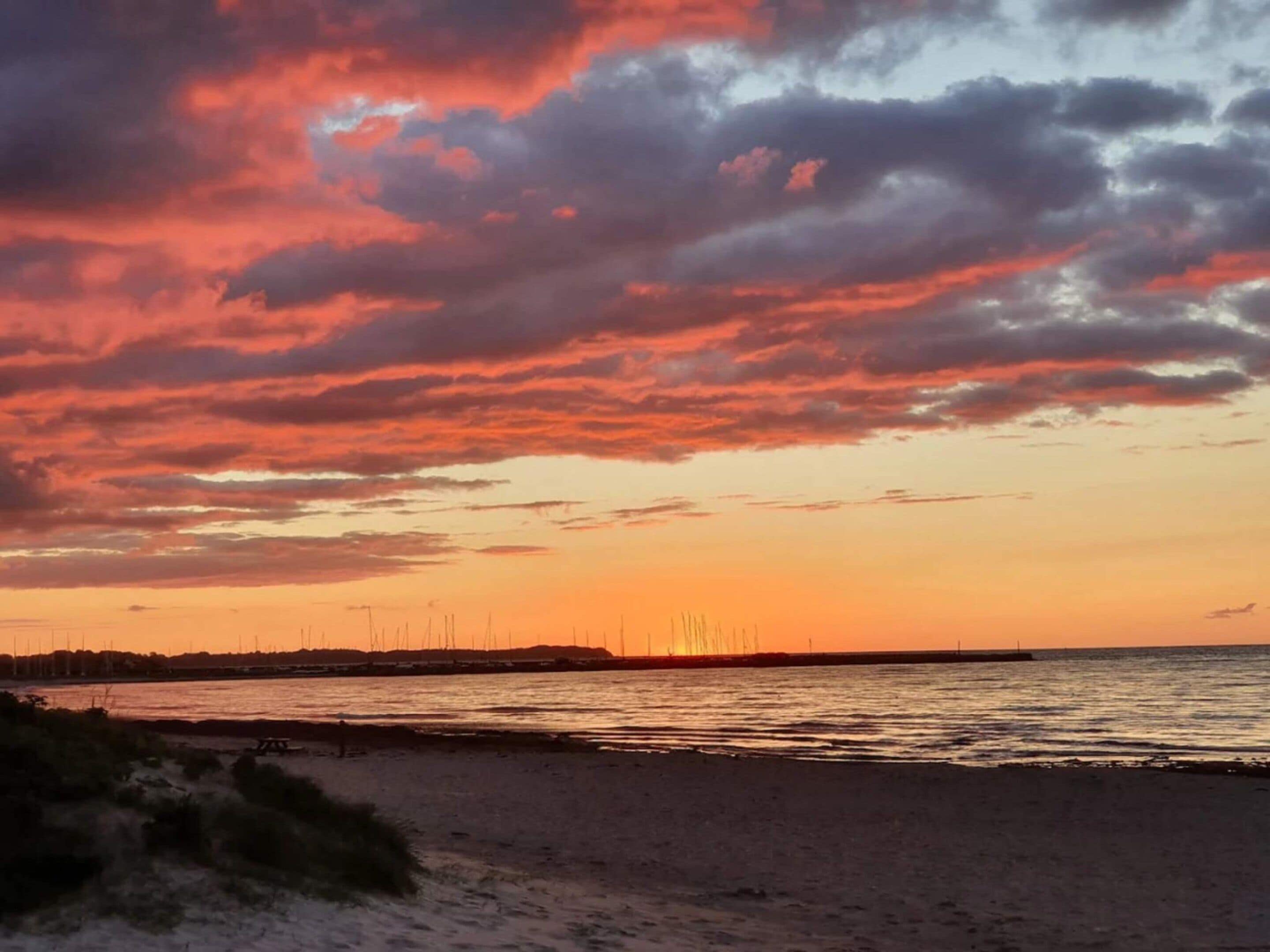 Uma praia com um pôr do sol, um píer ao longe e um céu claro.