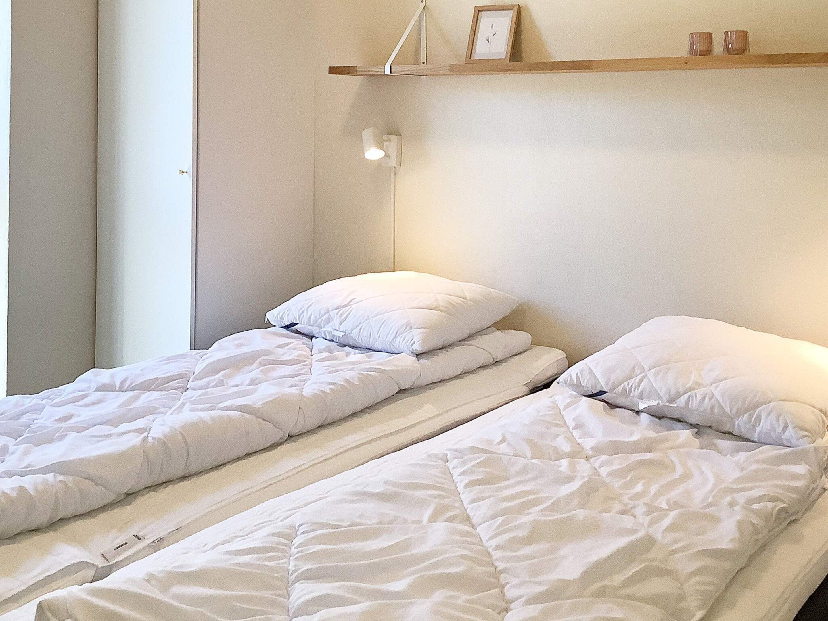 Quarto com duas camas de solteiro, cada uma com edredom e travesseiro brancos, uma mesinha de cabeceira com luminária e uma prateleira sobre as camas.