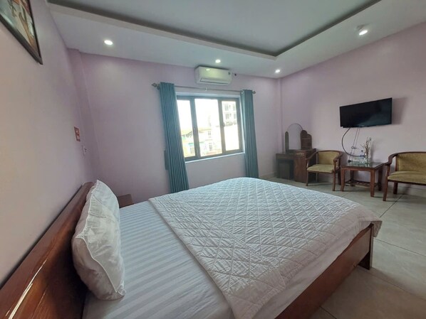 Quarto de hotel com cama, escrivaninha com cadeira, televisão e janela com cortinas.