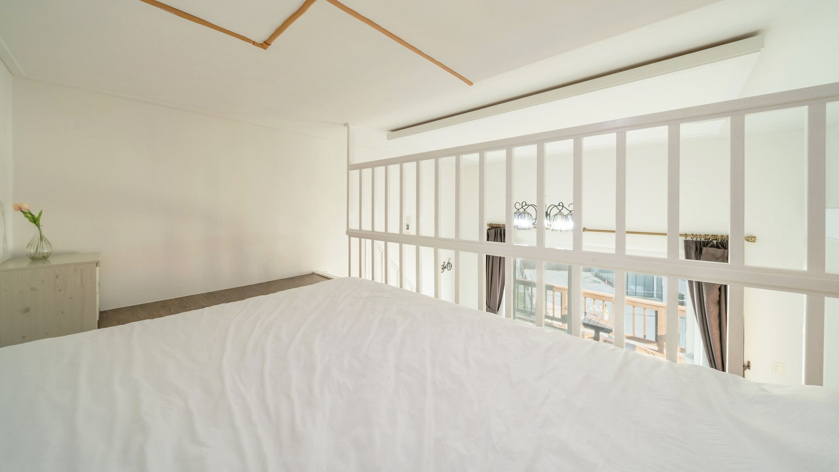 Quarto com uma cama grande, penteadeira, vaso com flores e vista para edifícios através de uma janela com guarda-corrimão branco.