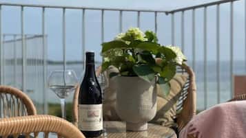 Uma cadeira de vime, um copo de vinho, uma garrafa de vinho e um vaso com planta em uma varanda com vista para o mar.