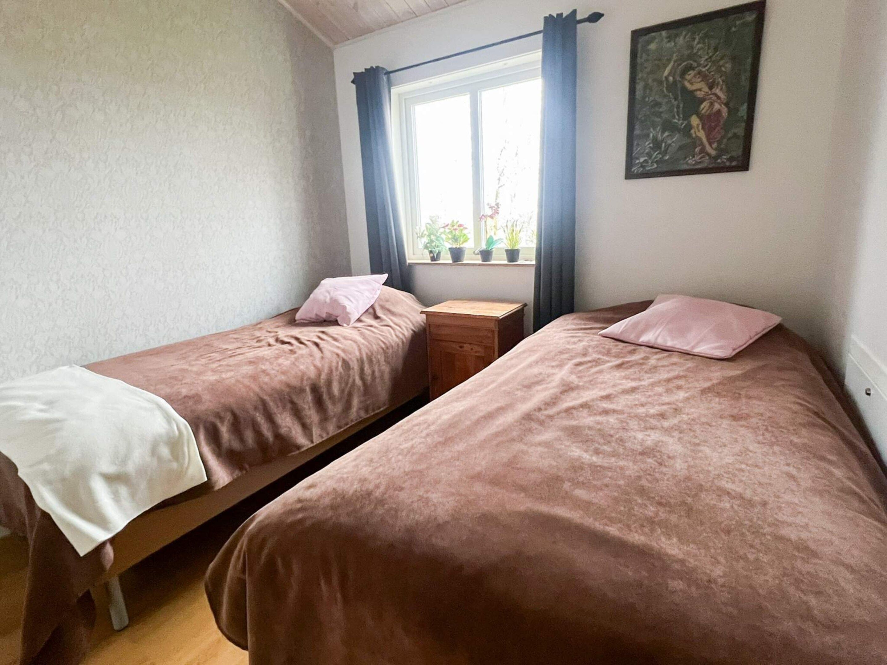 Quarto com duas camas de solteiro, uma pequena mesa de cabeceira de madeira, uma janela com cortinas e um quadro na parede.