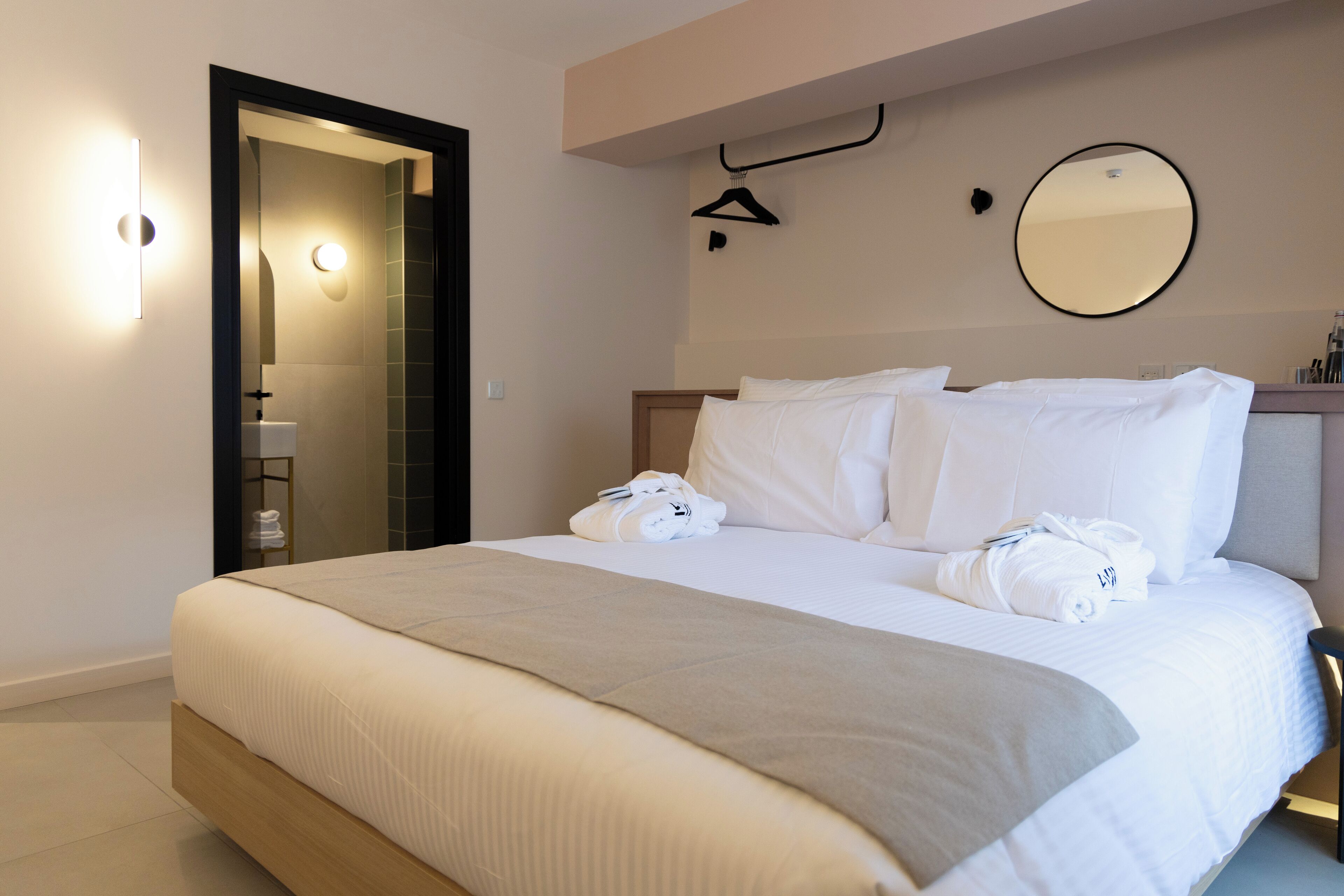 Quarto de hotel com uma cama grande, uma mesa de cabeceira, um espelho e um porta-toalhas fixado na parede.