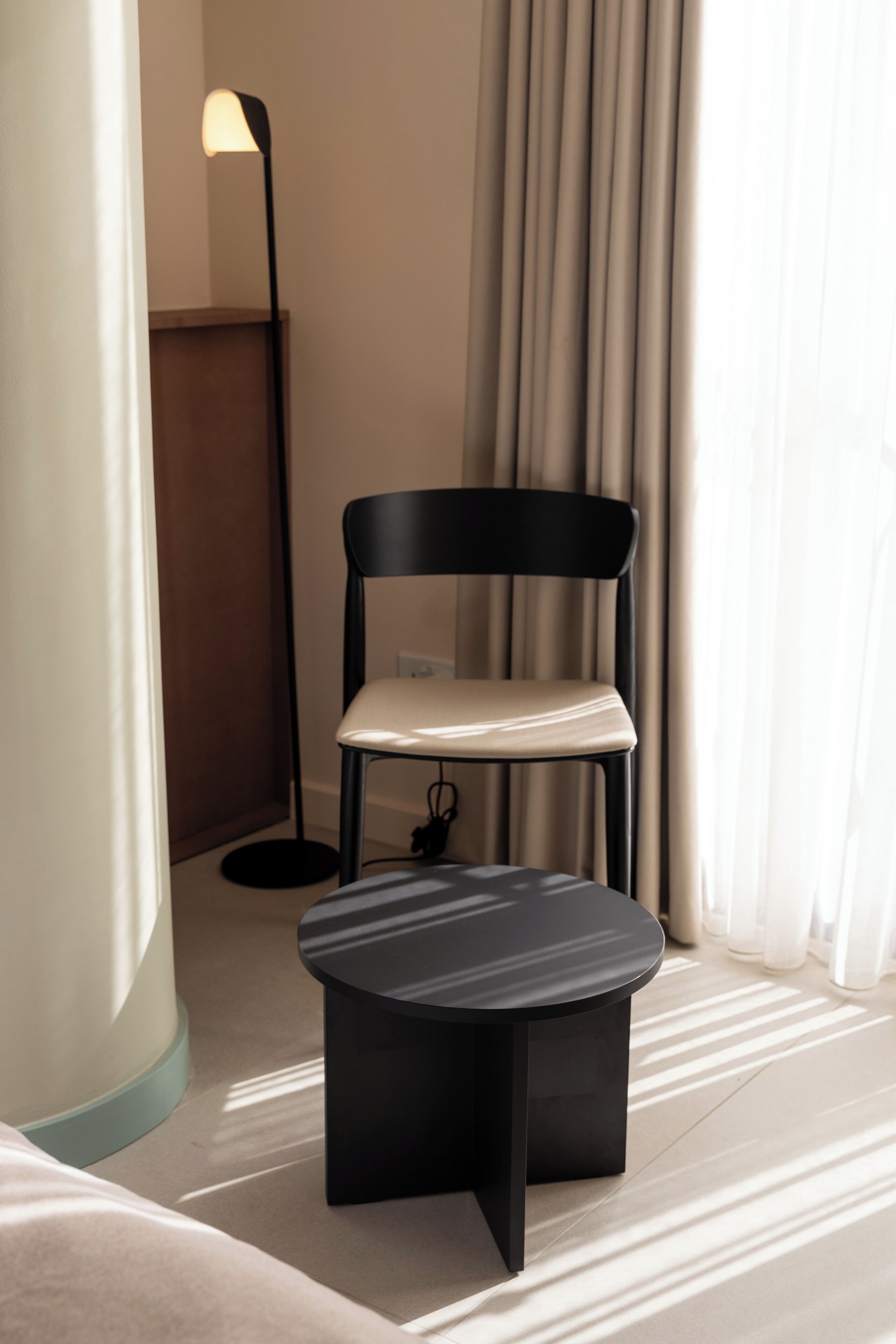 Quarto moderno com uma cadeira preta, uma mesa pequena, um abajur e uma janela com cortinas.