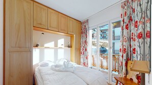 Quarto com cama, armários de madeira, escrivaninha e vista para uma varanda com grades.