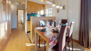Uma sala de jantar com uma mesa de madeira posta para quatro pessoas, uma cozinha ao fundo e vista para o exterior.