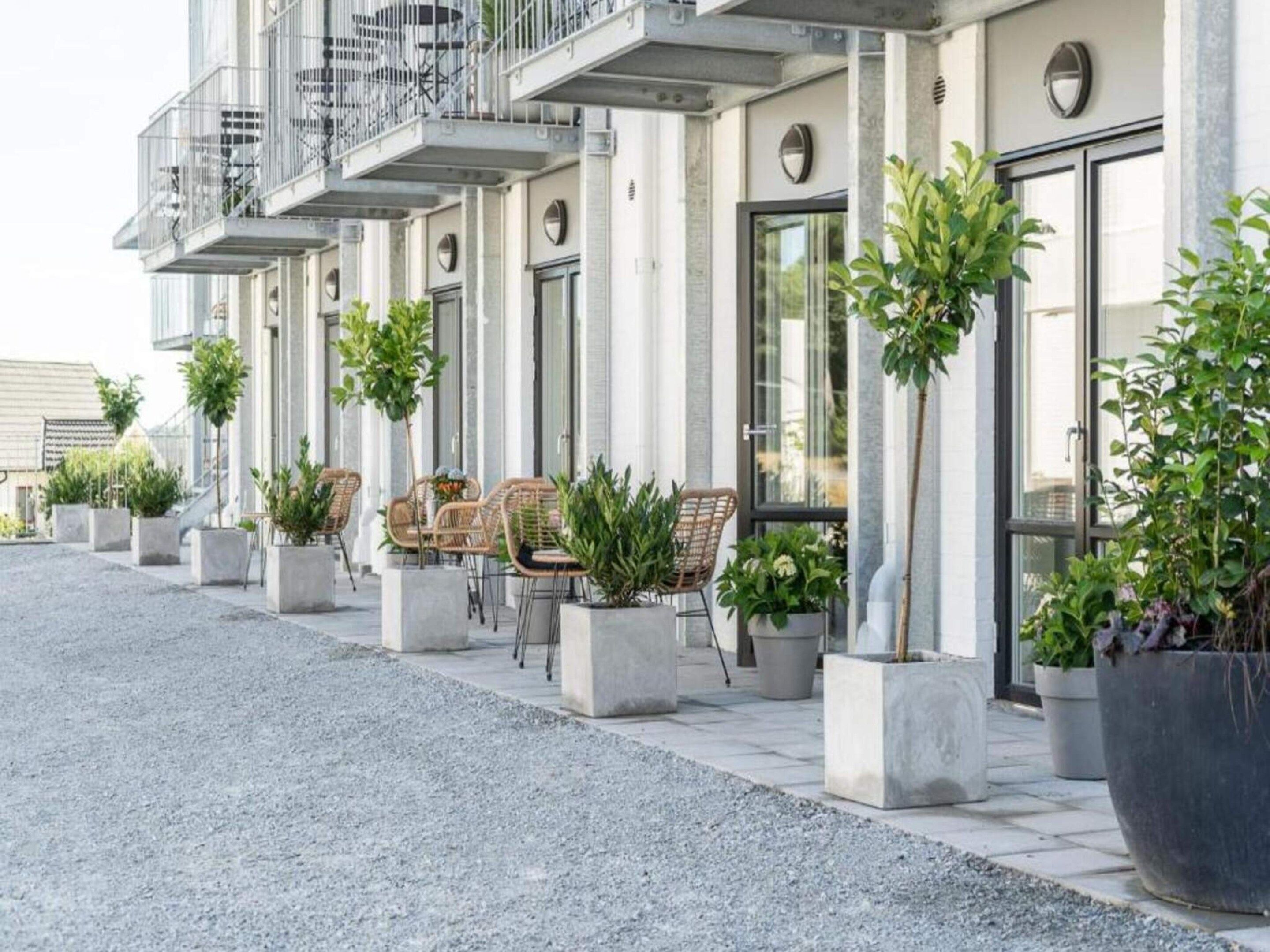 Um edifício com arquitetura moderna, apresentando uma série de varandas com guarda-corpos de metal, uma fileira de vasos com plantas e área de estar externa com cadeiras de vime.