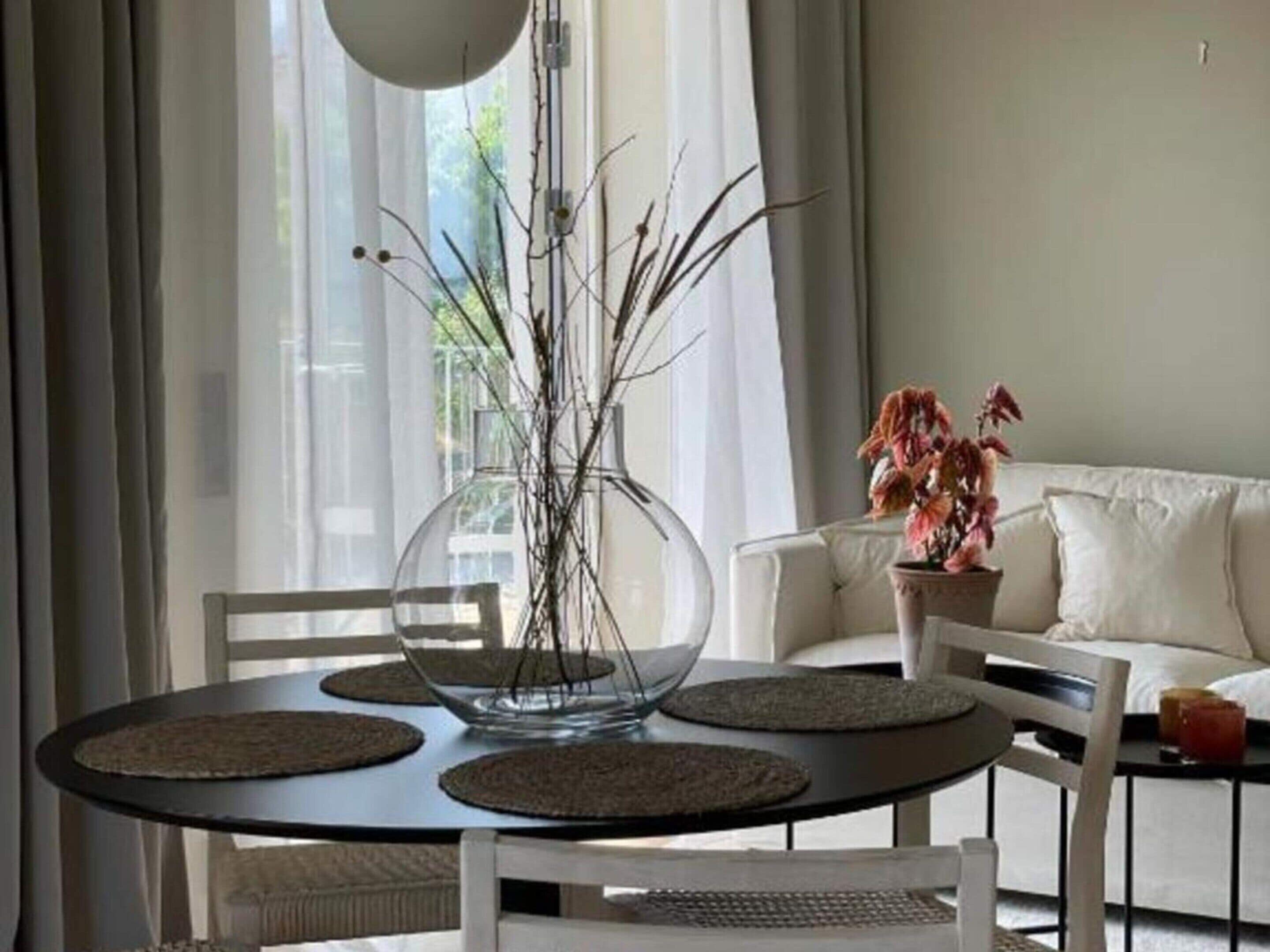 Uma área de refeições com uma mesa redonda, cadeiras brancas e um vaso com ramos.