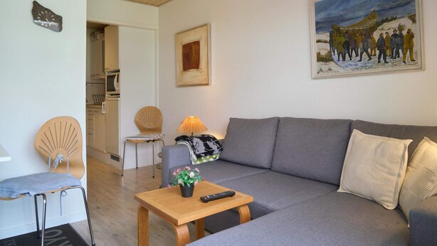 Uma sala de estar com um sofá cinzento, uma mesa de centro de madeira com um controle remoto, um abajur e um quadro de uma paisagem de inverno.