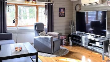 Uma sala de estar com televisão, uma poltrona cinza, um sofá, uma mesa de centro e uma janela com cortinas.