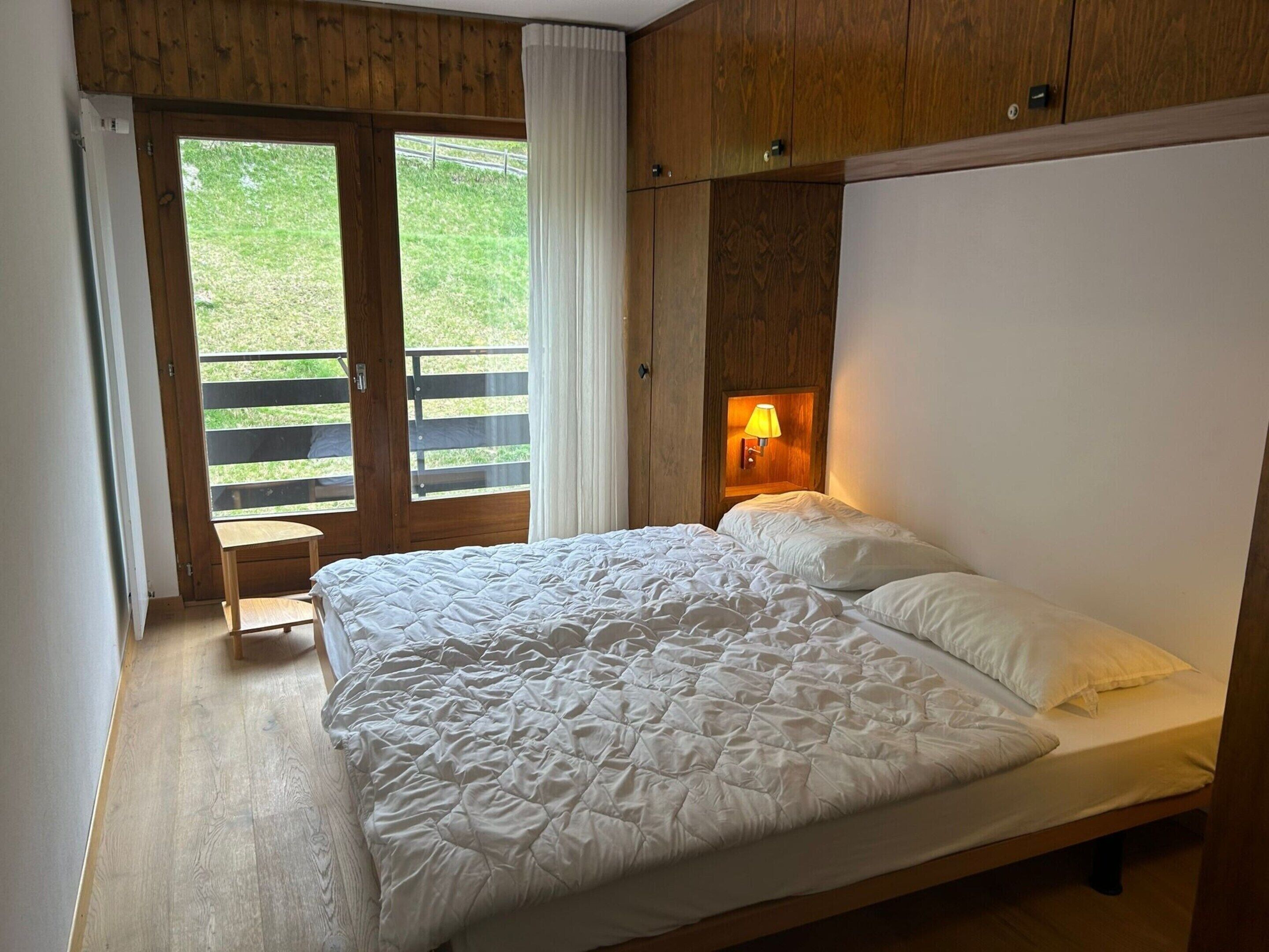 Quarto com estrutura de cama de madeira, luminária de cabeceira e vista para uma varanda com corrimão de madeira.