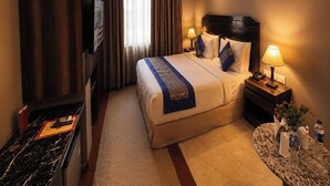 Quarto de hotel com cama, luminárias de cabeceira, ventoinha no teto, televisão e uma mesinha com um vaso e garrafas de água.