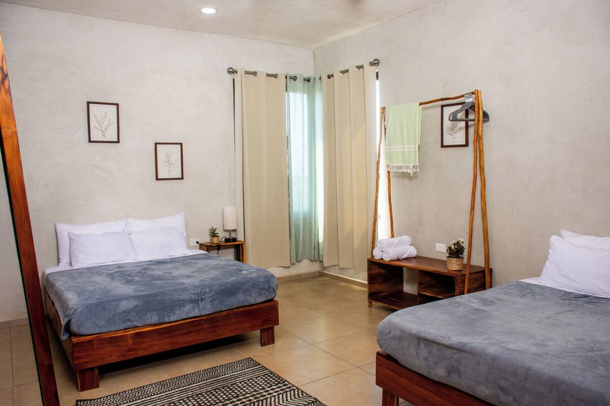 Quarto de hotel com duas camas de madeira, uma mesinha de cabeceira com abajur, um quadro emoldurado, um cortina de chuveiro e um porta-toalhas.