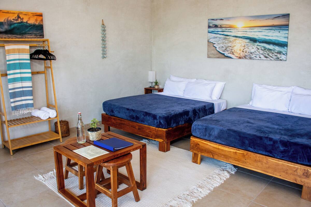 Quarto com duas camas, uma mesa de madeira, um banquinho pequeno, uma prateleira com toalhas e uma pintura de uma cena de praia.