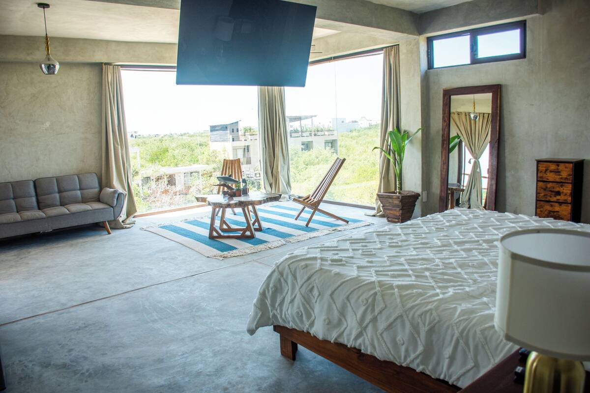 Quarto com cama grande, sofá, televisão e vista para área verde.