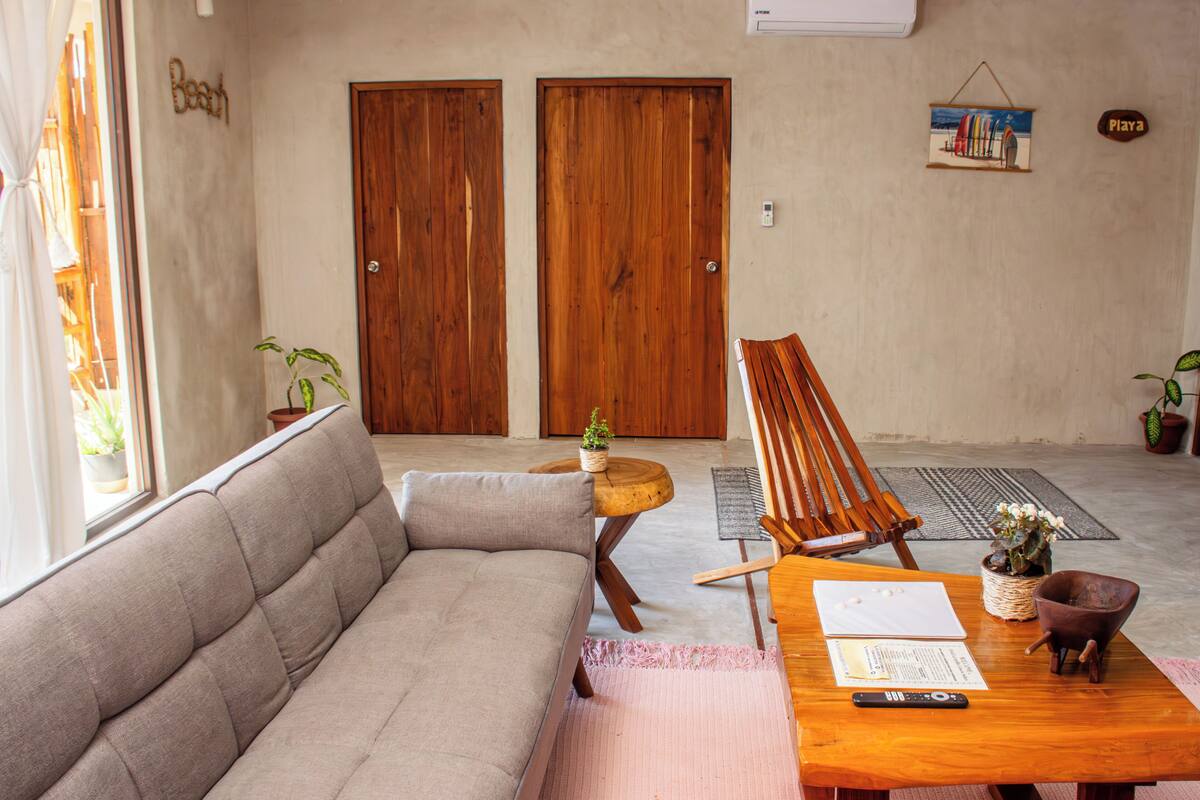 Quarto com sofá, mesa de madeira com papéis e um controle remoto, duas portas de madeira e uma pequena planta no chão.