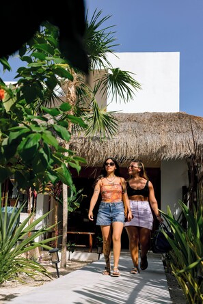 Duas pessoas caminhando por um caminho entre plantas tropicais e uma construção com telhado de palha.