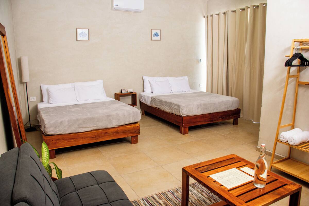 Quarto de hotel com duas camas de madeira, um sofá, uma mesa de madeira e uma estante com toalhas.