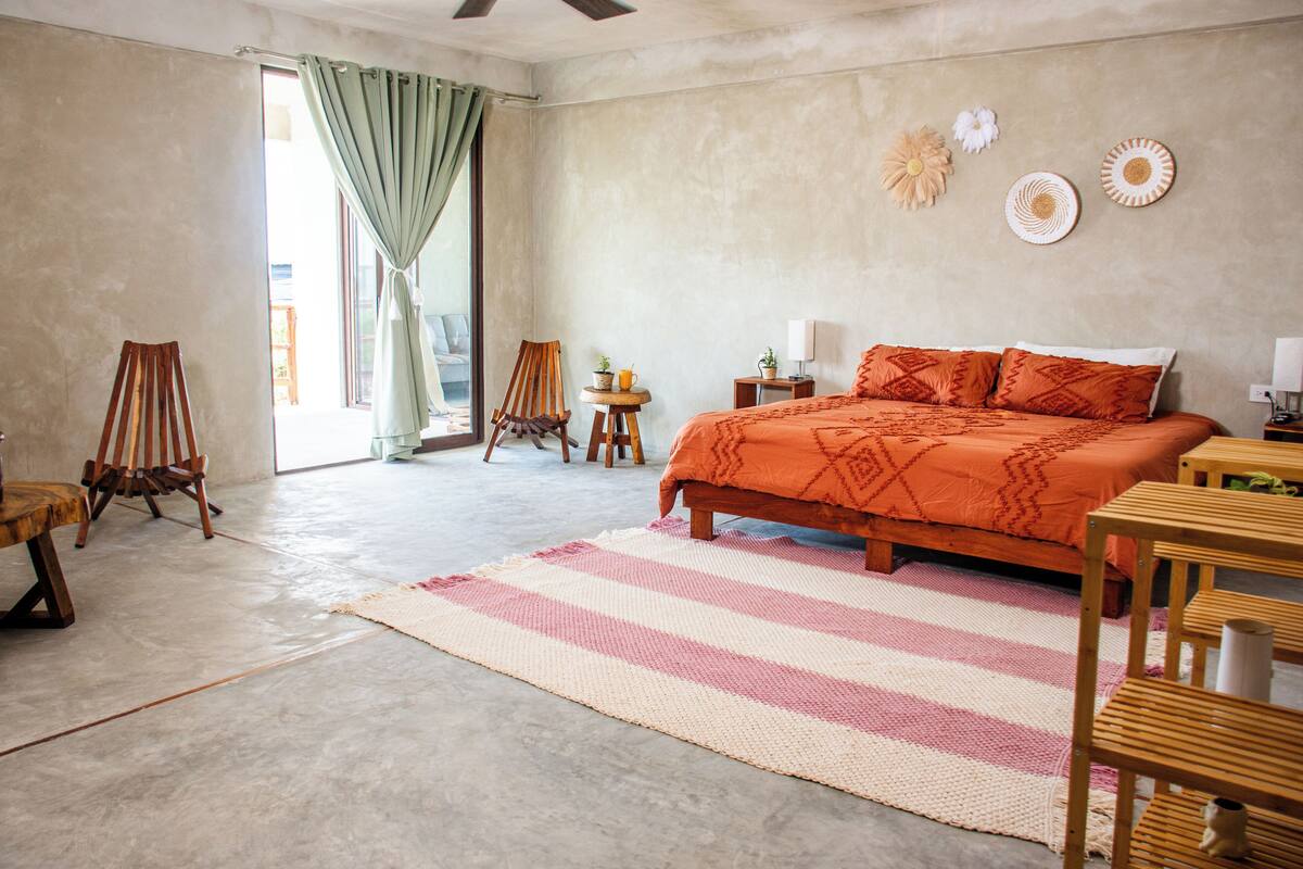 Um quarto com uma cama, uma cadeira de madeira, uma mesinha, um abajur e uma janela com cortinas.