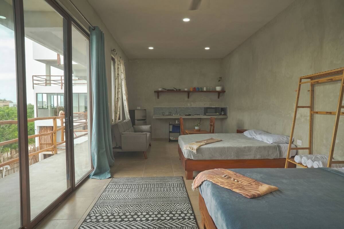 Quarto de hotel moderno com duas camas, varanda com vista, cozinha compacta e área de estar com sofá e mesa.