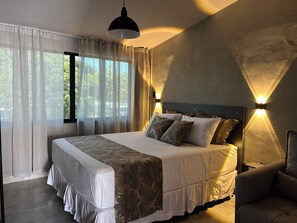 Quarto moderno com uma cama grande, cabeceira com iluminação integrada, cortinas leves e vista para o verde lá fora.