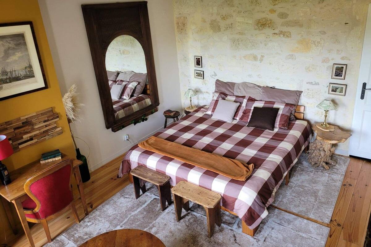 Quarto com colcha xadrez, um banco de madeira, uma escrivaninha com cadeira, um espelho e uma parede rústica.