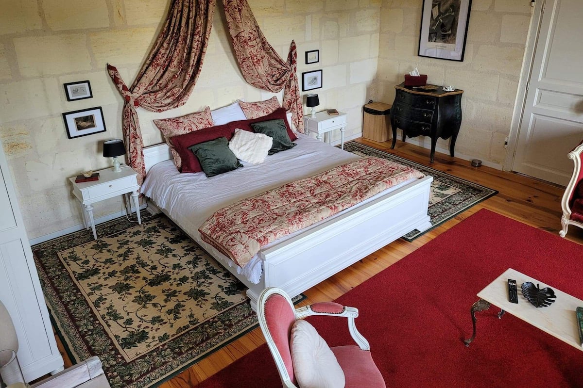Quarto com uma cama grande, um tapete vermelho, piso de madeira, mesa de cabeceira, cômoda, cadeira e um quadro na parede.