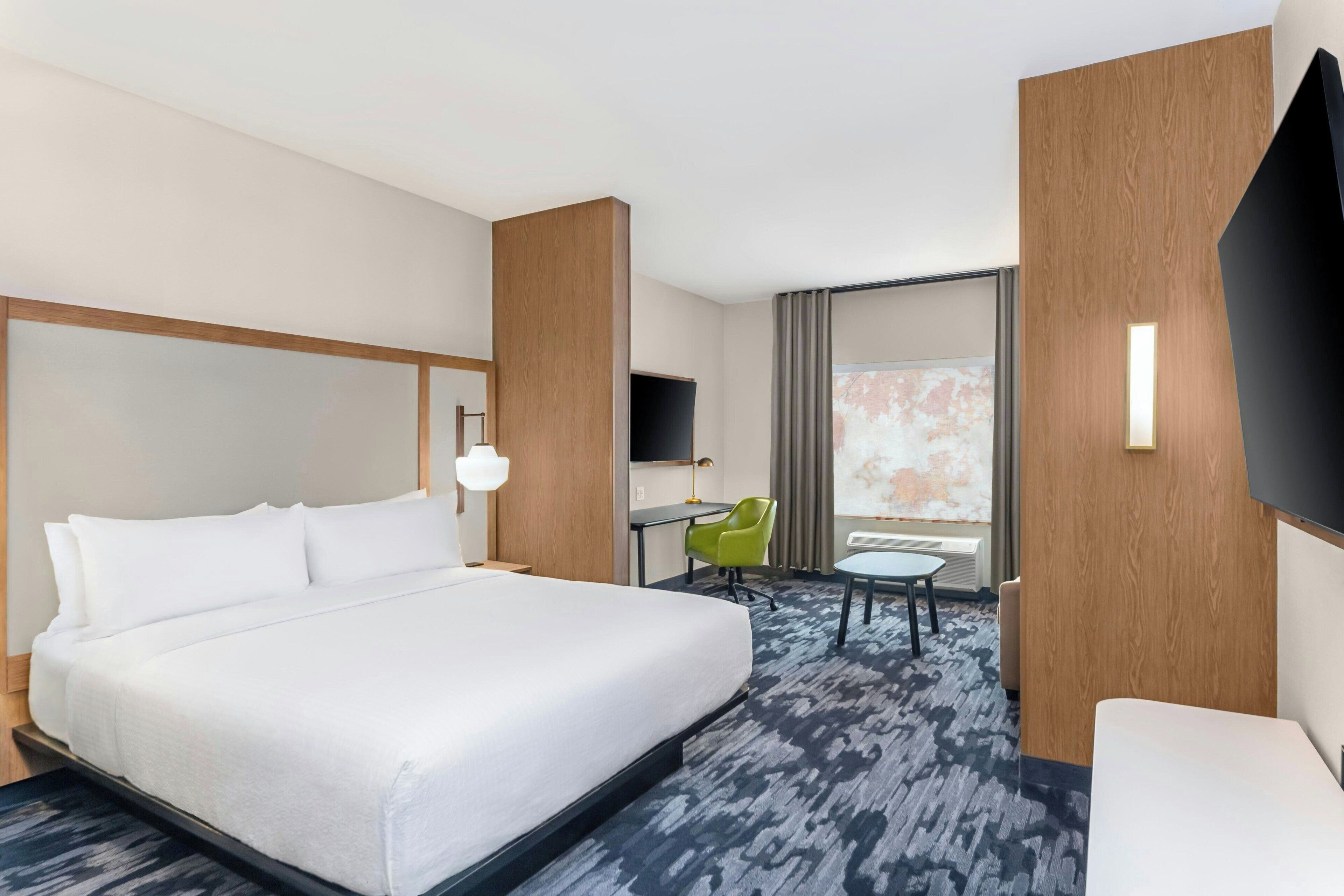 Quarto de hotel com uma cama grande, uma escrivaninha, uma cadeira, uma TV e uma mesinha.