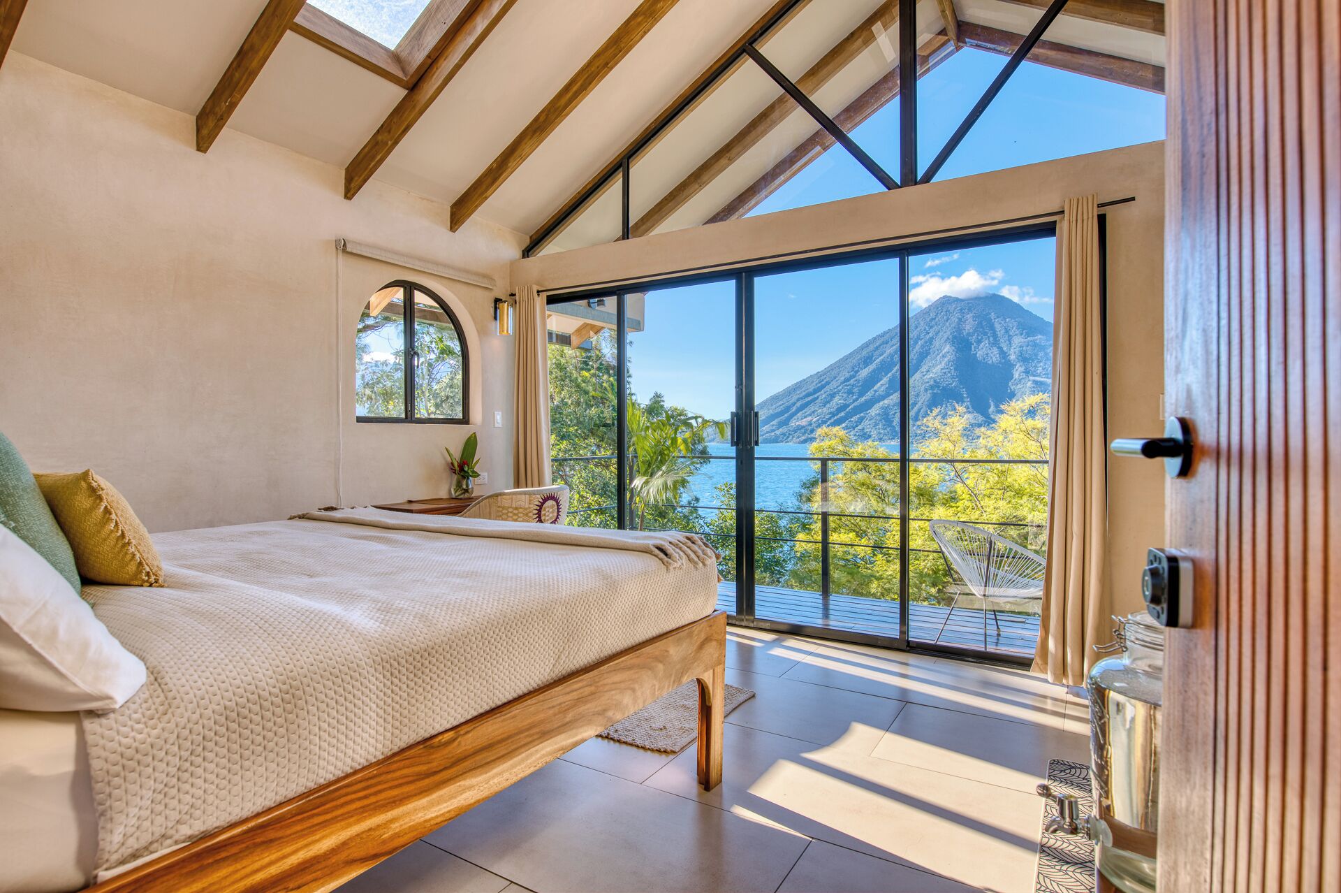 Quarto com cama grande, janela com vista para a montanha e mar, e estrutura de teto em madeira.