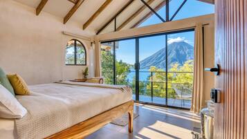 Quarto com cama grande, janela com vista para a montanha e mar, e estrutura de teto em madeira.