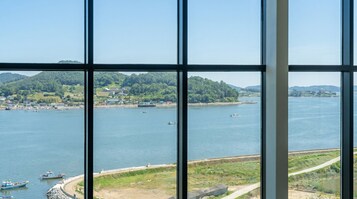 Vista pela janela de uma área costeira, com barcos, um cais e uma colina arborizada ao fundo.