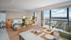 Quarto de hotel moderno com uma cama grande, televisão, área de jantar com mesa posta para dois e vista para a cidade através de grandes janelas.