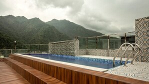 Piscina no terraço com deck de madeira, azulejos estampados e montanhas ao fundo.