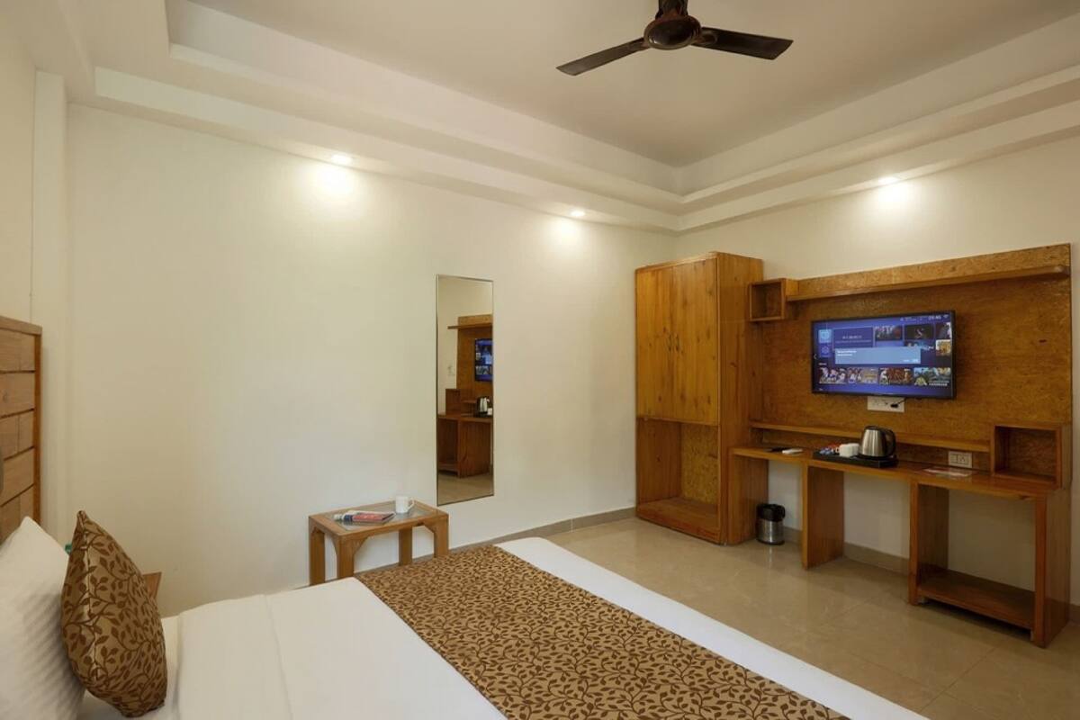 Quarto com cama, guarda-roupa de madeira, televisão na parede, uma pequena mesa com uma chávena e um ventilador de teto.