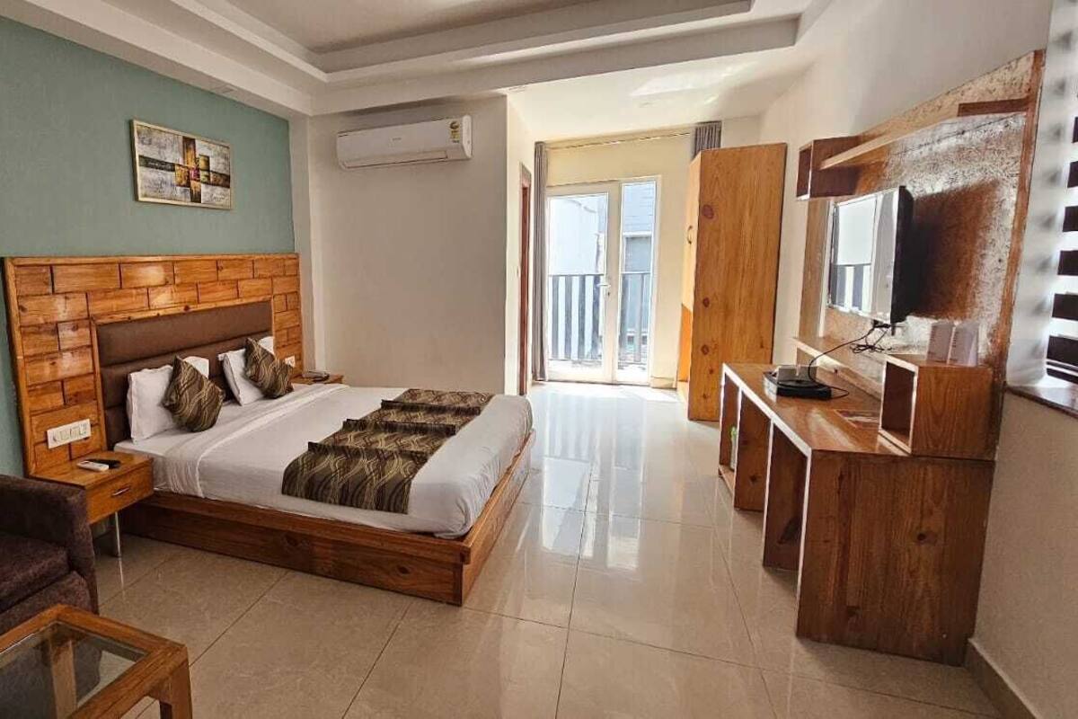 Quarto com cabeceira de madeira, cama com várias almofadas, cômoda de madeira, televisão e varanda visível através de uma porta aberta.