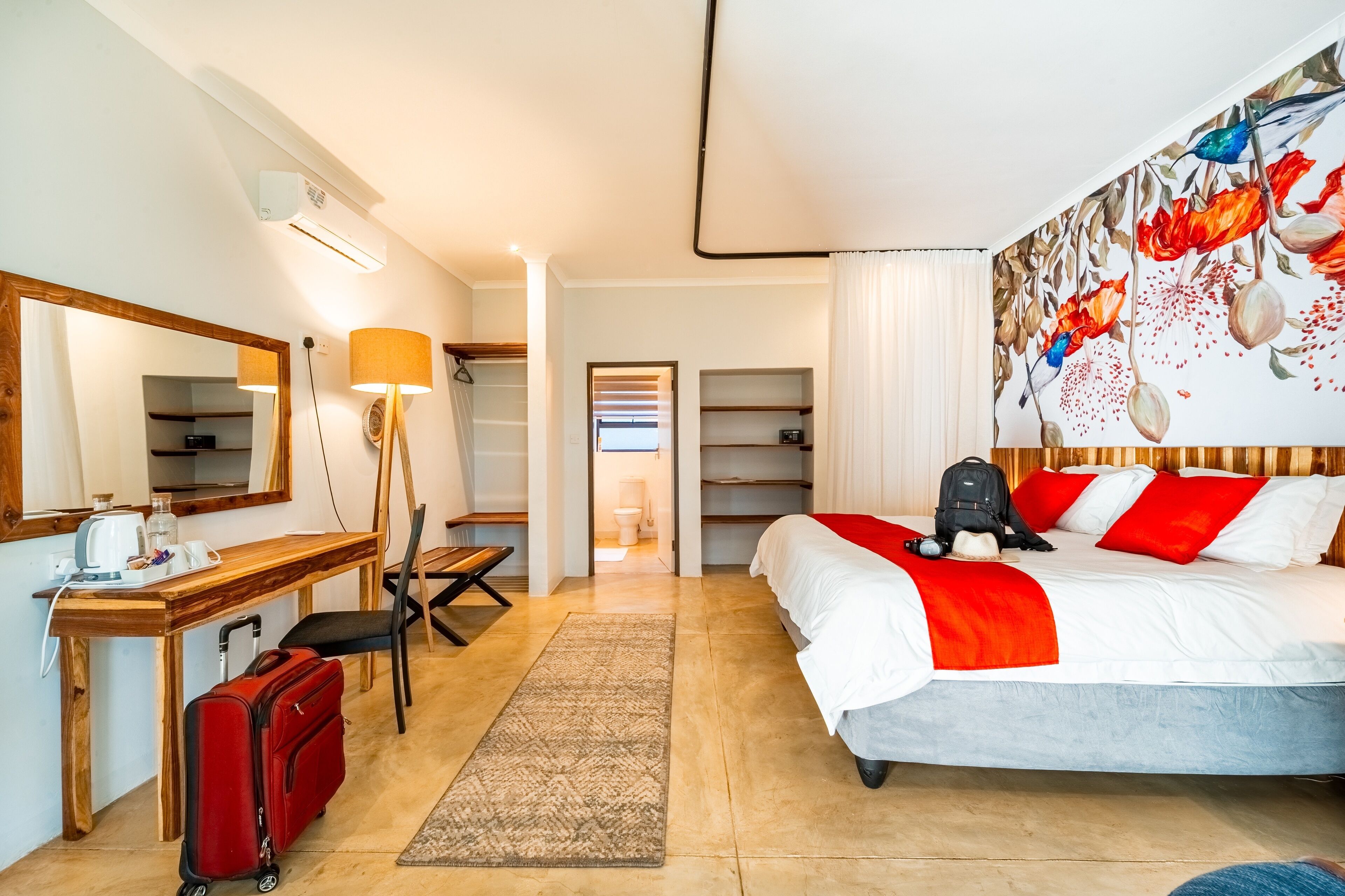 Quarto de hotel com cama, escrivaninha, cadeira, mala, luminária e um quadro na parede.