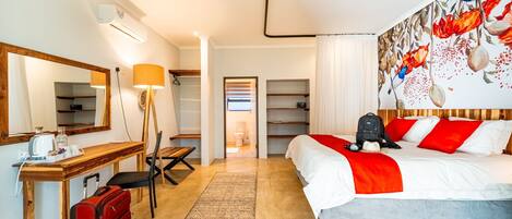 Quarto de hotel com cama, escrivaninha, cadeira, mala, luminária e um quadro na parede.