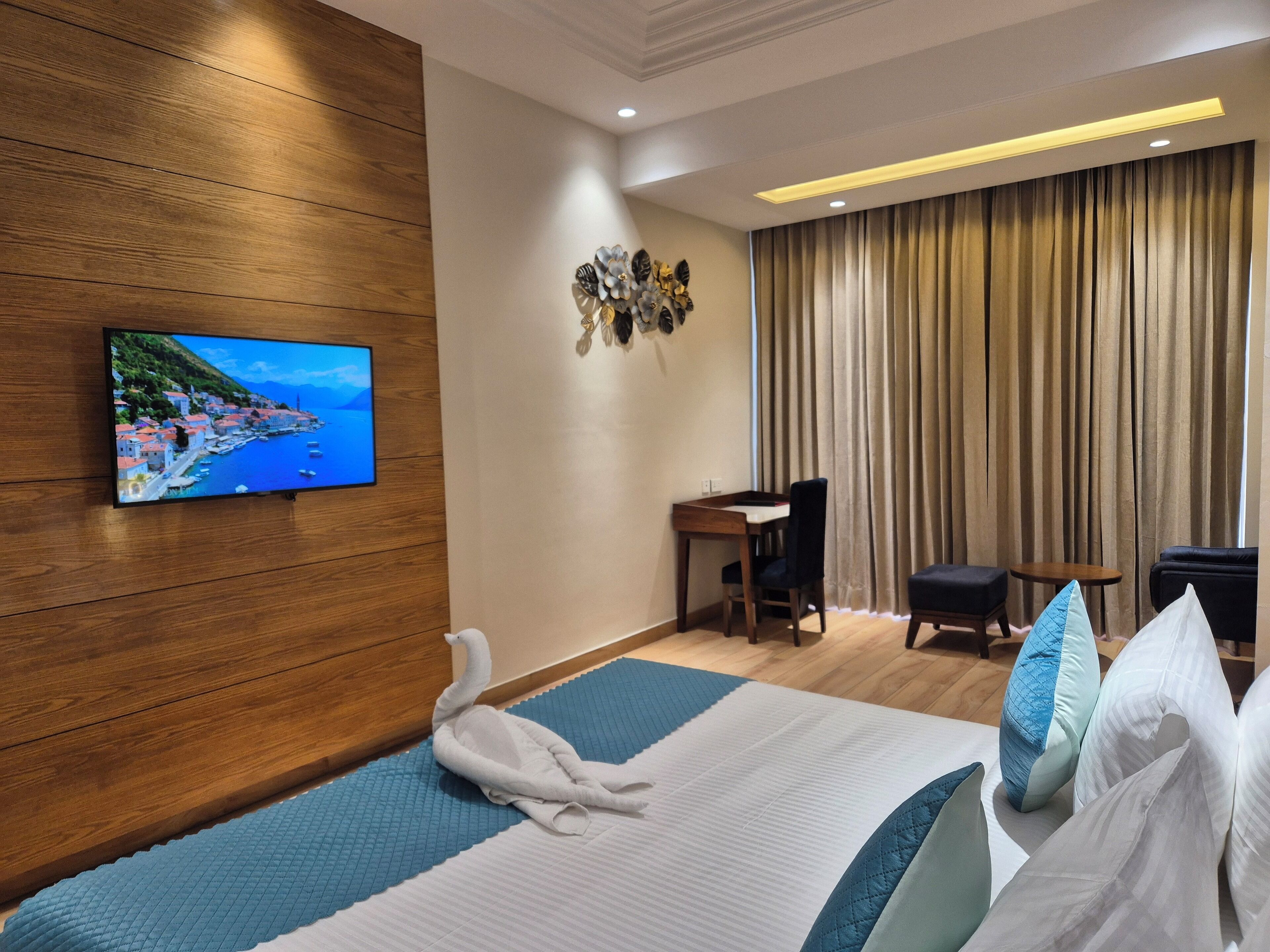 Quarto de hotel com parede de painel de madeira, uma televisão exibindo uma vista panorâmica, uma escrivaninha com cadeira, uma cama com roupa de cama azul e branca, e uma janela grande com cortinas.