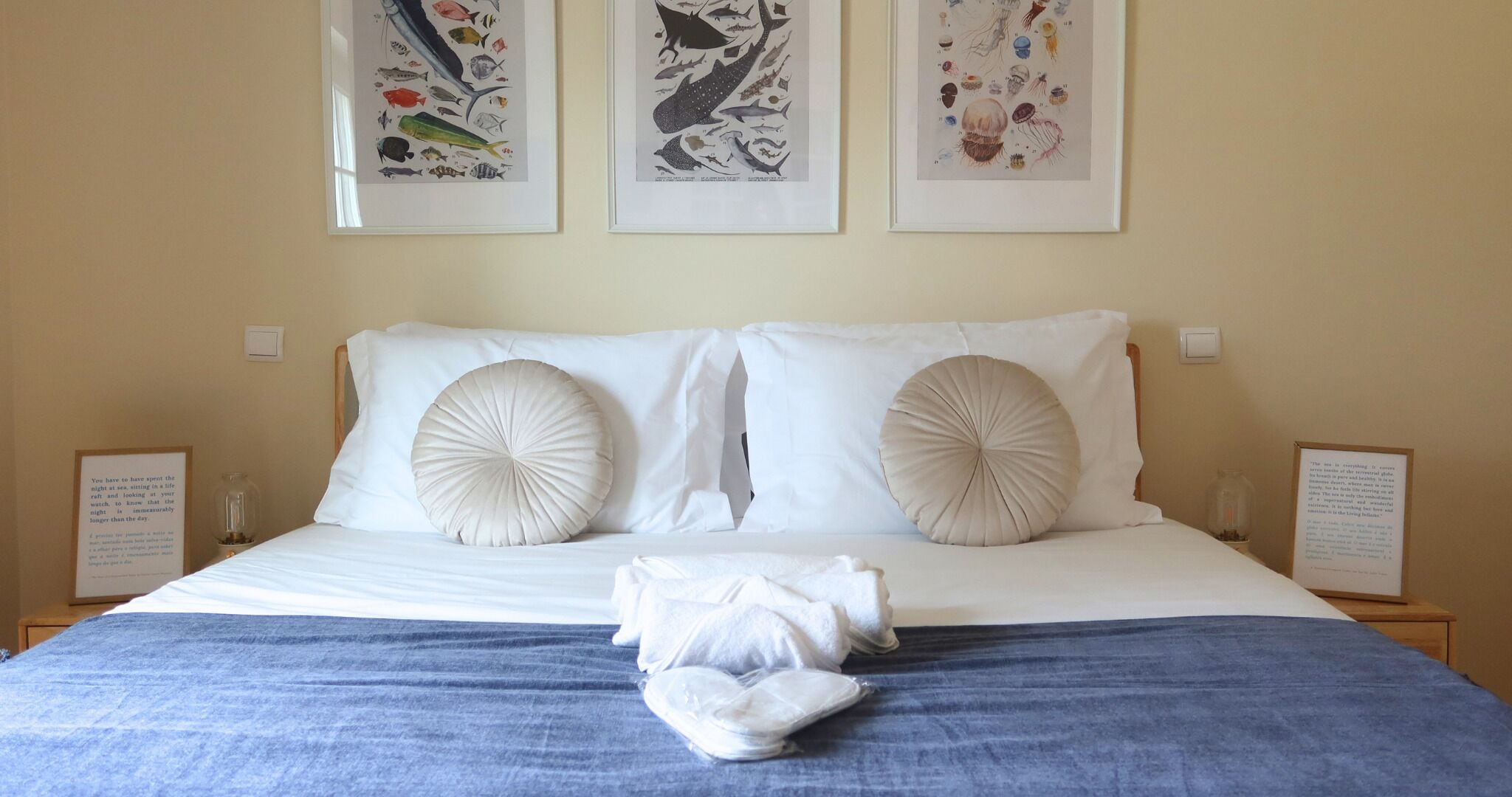 Uma cama bem arrumada com lençóis brancos, duas almofadas decorativas grandes e uma toalha dobrada sobre a cama. Há três quadros na parede acima da cama.