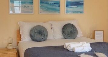 Uma cama bem arrumada com duas almofadas, uma mesa de cabeceira com abajur, e três obras de arte emolduradas na parede.