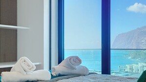 Quarto de hotel com uma janela ampla que oferece vista para o mar e um penhasco. Há toalhas na cama e uma estante com um painel listrado.