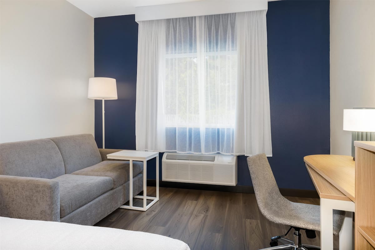 Quarto de hotel com sofá cinzento, uma mesa pequena, uma cadeira, uma escrivaninha e uma janela com cortinas leves.