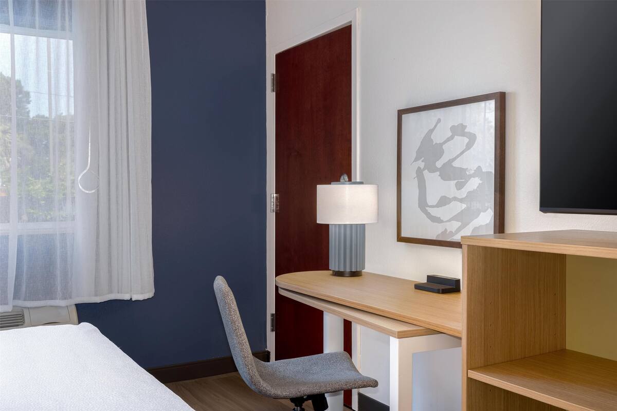 Quarto de hotel com cama, escrivaninha com cadeira, luminária e uma obra de arte emoldurada na parede.