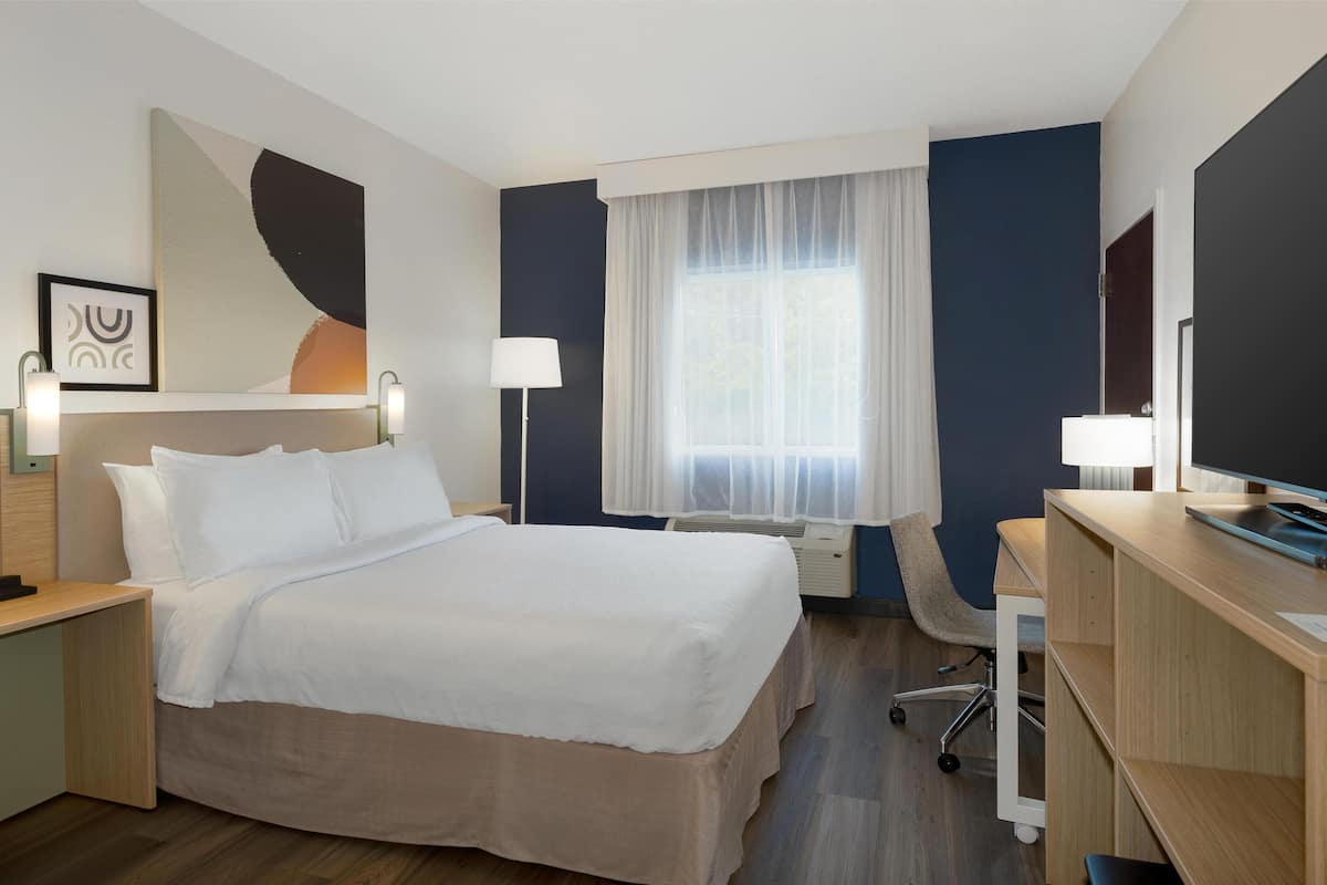 Quarto de hotel com cama, escrivaninha, cadeira e televisão.