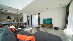 Uma sala de estar moderna com um sofá cinzento, almofadas laranja, uma TV e um espaço de jantar com mesa e cadeiras de madeira.